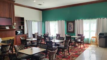 Um restaurante com painéis de madeira, paredes verdes e cortinas brancas. Mesas e cadeiras dispostas para refeições.