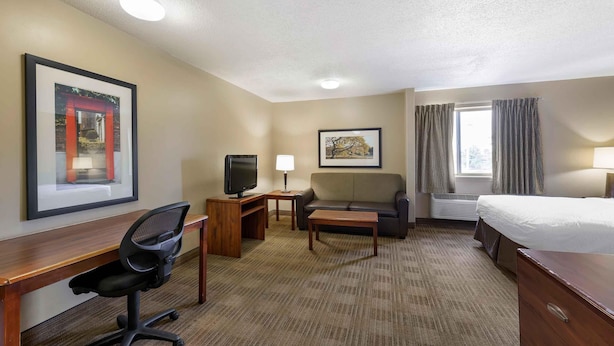 Quarto de hotel com cama, escrivaninha, cadeira, televisão e duas fotografias emolduradas nas paredes.