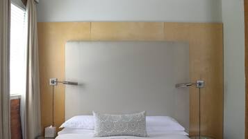 Uma cama grande com lençóis brancos, cabeceira de madeira, abajures e uma janela com cortinas.
