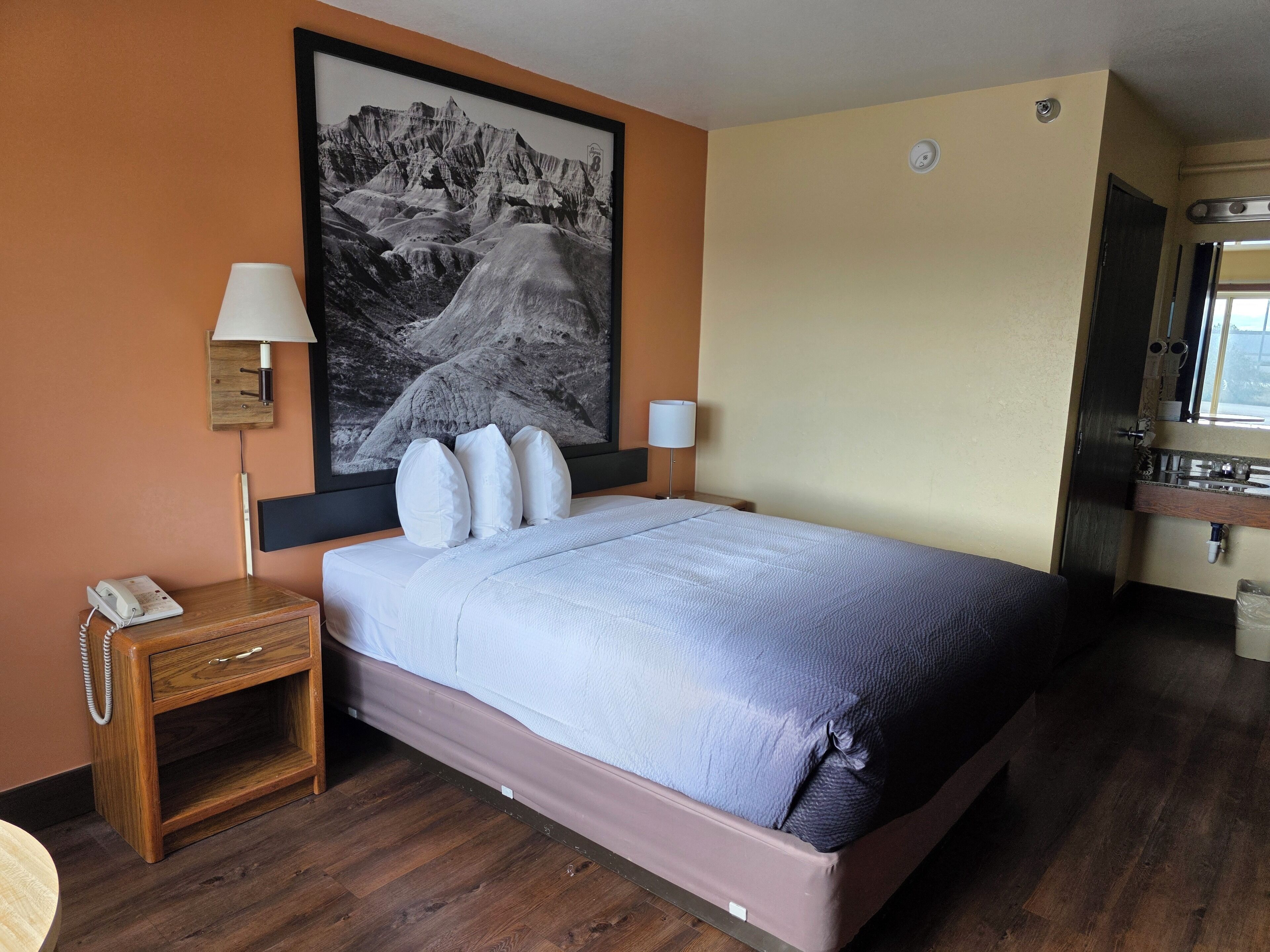 Quarto de hotel com cama, mesa de cabeceira, abajur, pintura e banheiro visível por uma porta aberta.