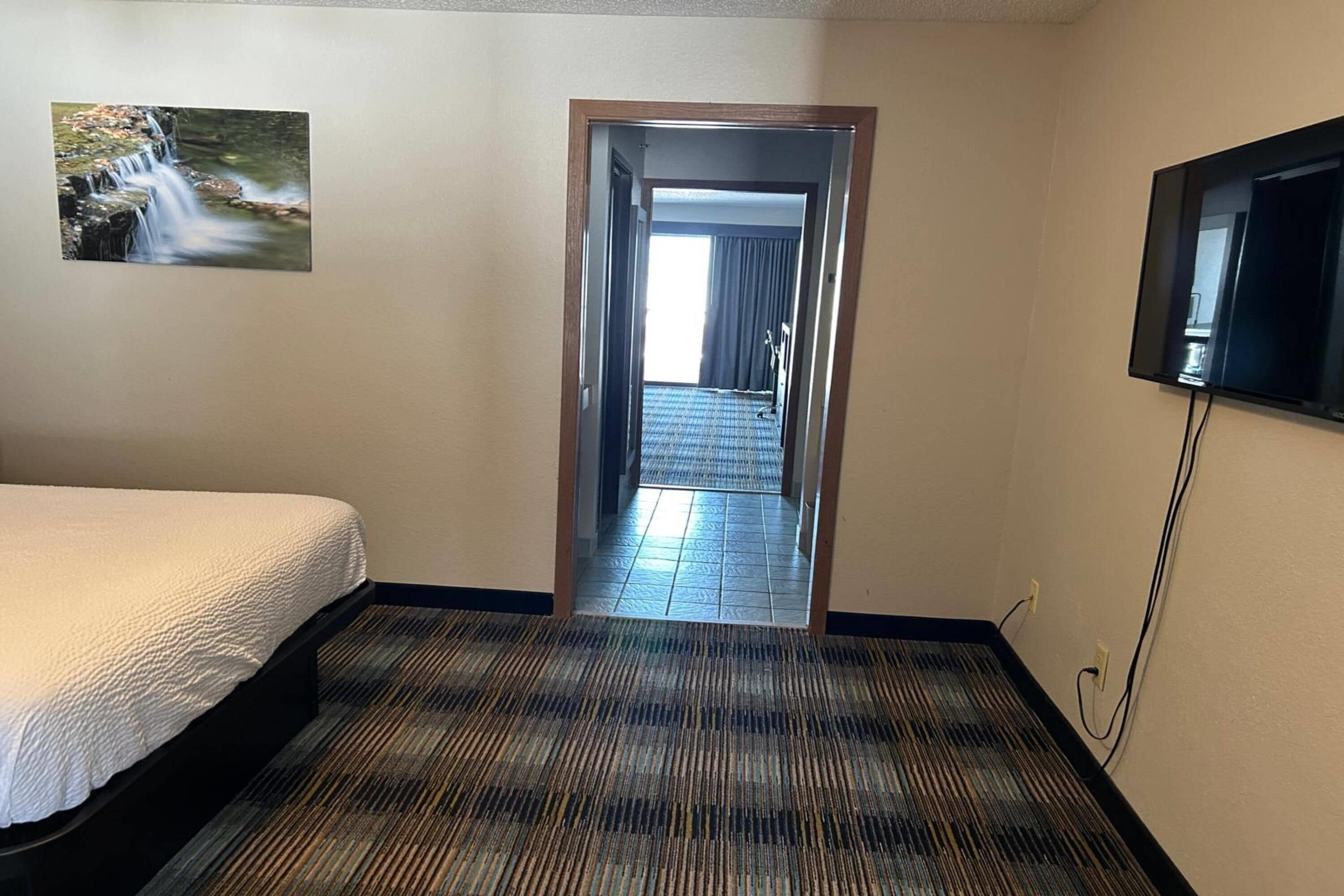 Quarto de hotel com cama, piso coberto por carpete, televisão e porta que dá para outro cômodo.