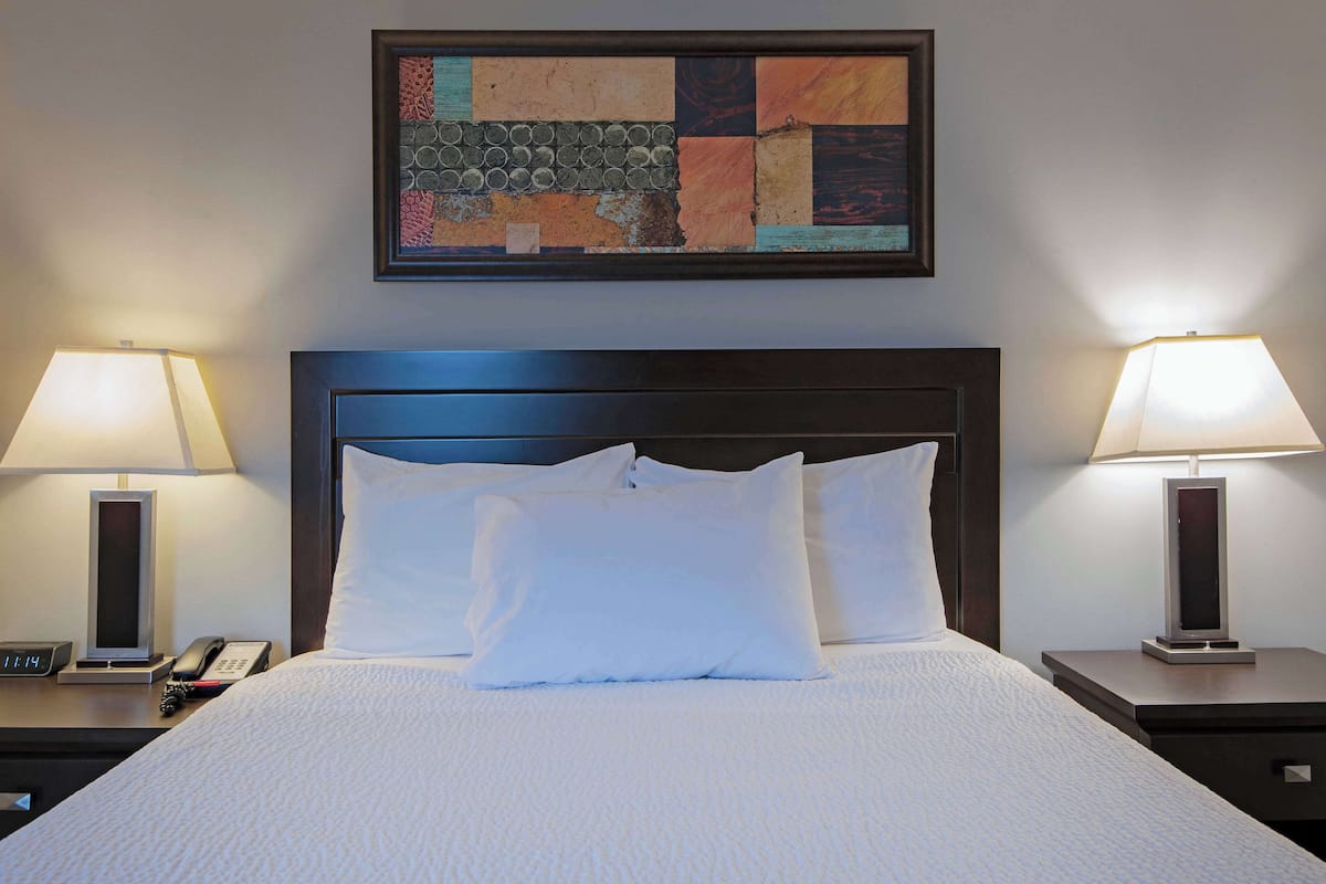 Uma cama bem arrumada com lençóis brancos, duas luminárias de cabeceira, um relógio digital e uma obra de arte emoldurada acima da cama.