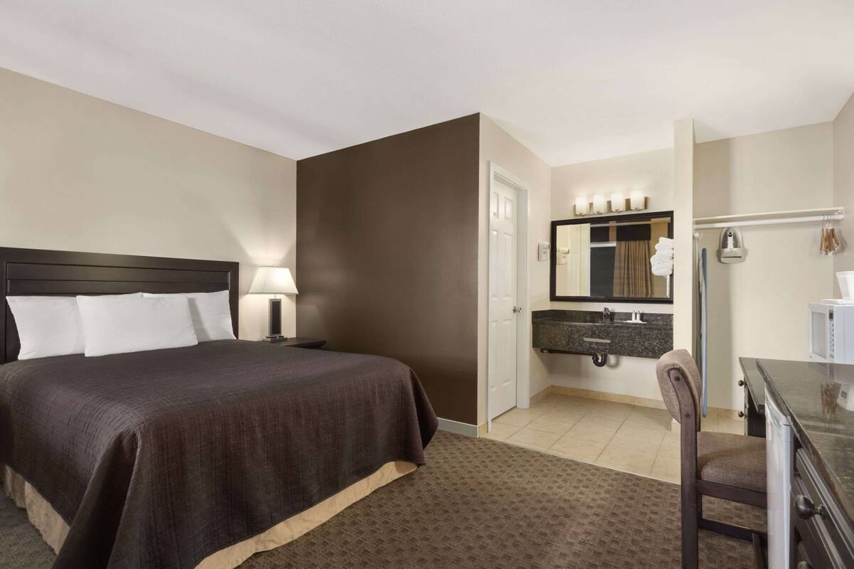 Quarto de hotel com uma cama grande, uma mesinha de cabeceira com abajur, banheiro com pia e espelho, e uma cadeira.