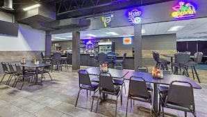 Restaurante com várias mesas e cadeiras, área de bar e letreiros de neon das marcas de cerveja Corona e Modelo.