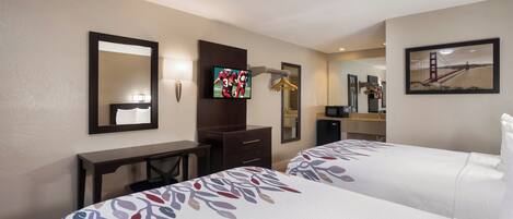 Quarto de hotel com duas camas, uma escrivaninha, televisão, espelho, guarda-roupa e banheiro visível por uma porta aberta.