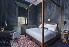Uma cama de dossel com estrutura de madeira e lençóis brancos, uma poltrona estofada, uma escrivaninha com abajur e um tapete estampado.