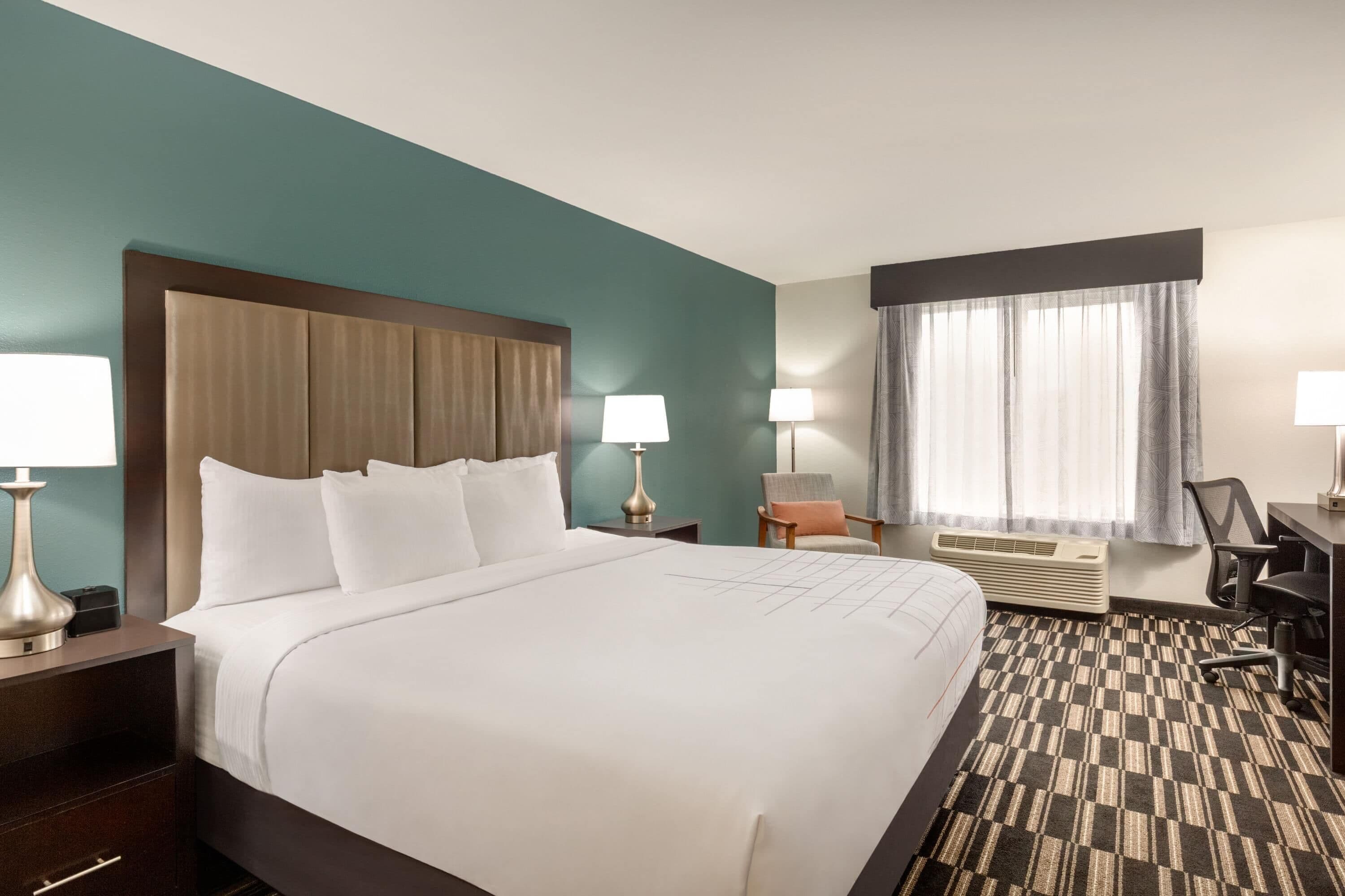 Quarto de hotel com uma cama grande, duas mesas de cabeceira com luminárias, uma escrivaninha com cadeira e uma janela com cortinas.