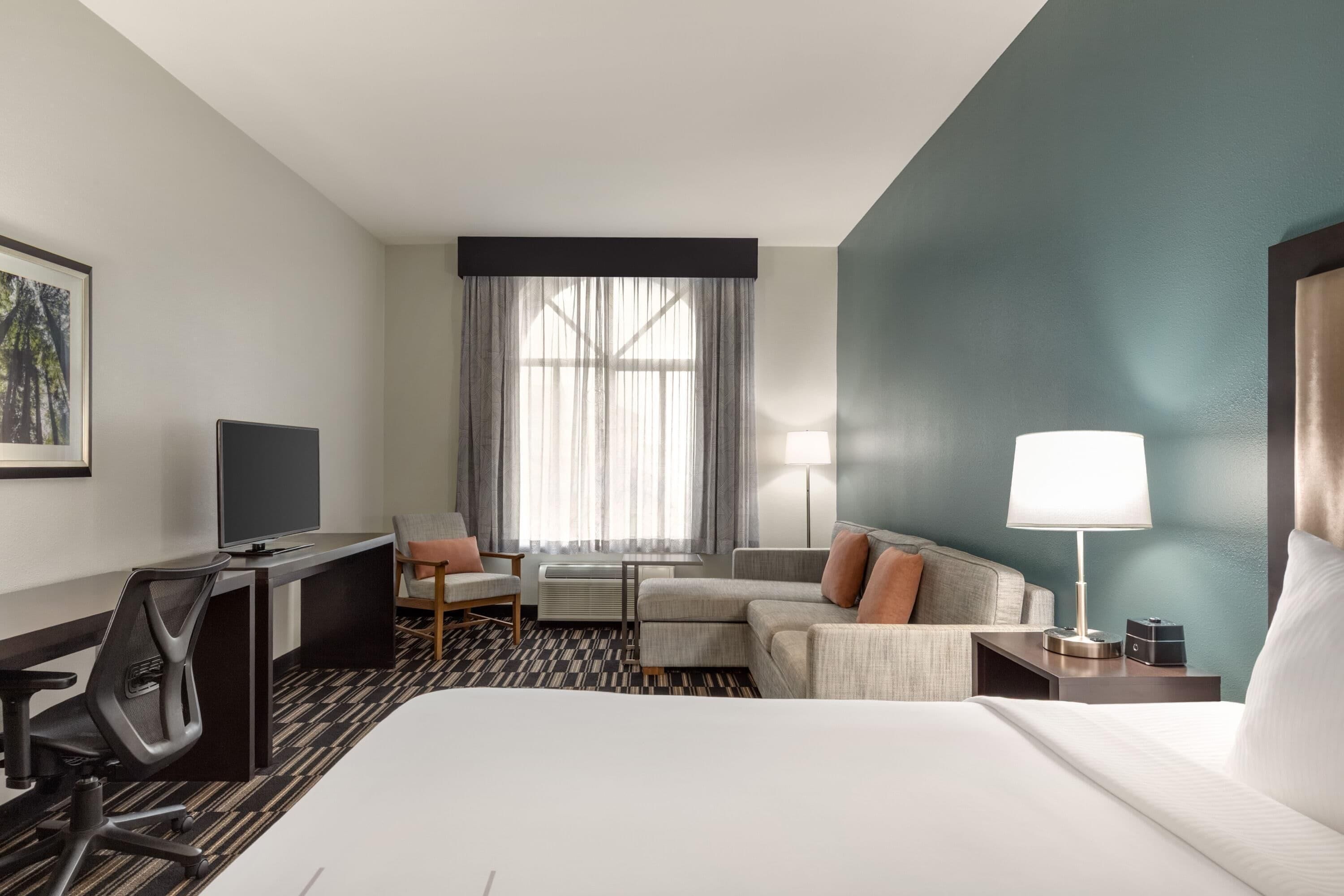 Quarto de hotel com cama, escrivaninha com computador, cadeira, sofá, luminária e janela com cortinas.