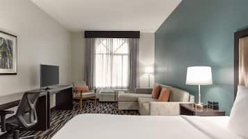 Quarto de hotel com cama, escrivaninha com computador, cadeira, sofá, luminária e janela com cortinas.