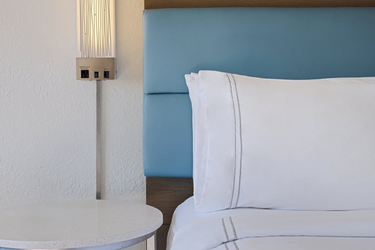 Quarto de hotel com uma mesa redonda, uma cama com roupa de cama branca e um candeeiro fixado na parede.
