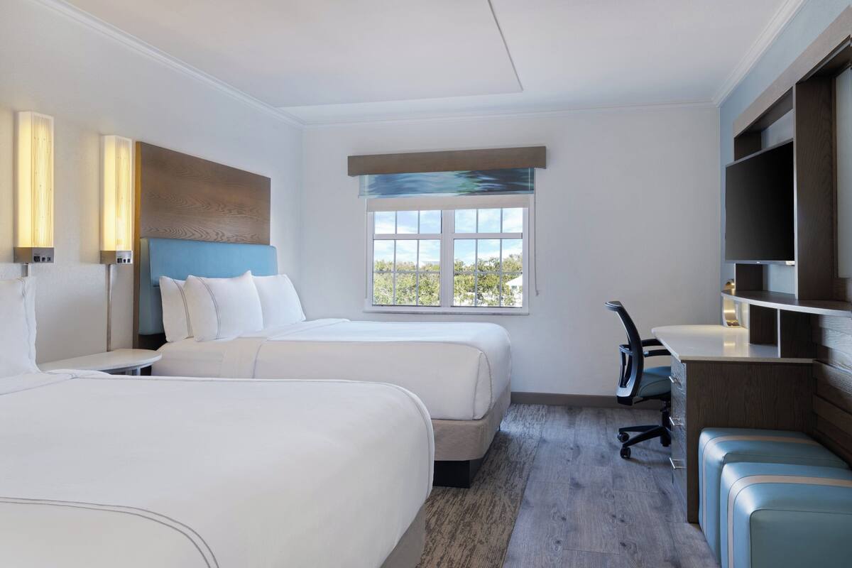Quarto de hotel com duas camas, uma escrivaninha, uma cadeira, uma televisão e uma janela com vista.