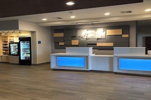 Uma área de recepção com design moderno, destacando um balcão com painéis azuis iluminados, uma placa fixada na parede e uma geladeira abastecida com bebidas.