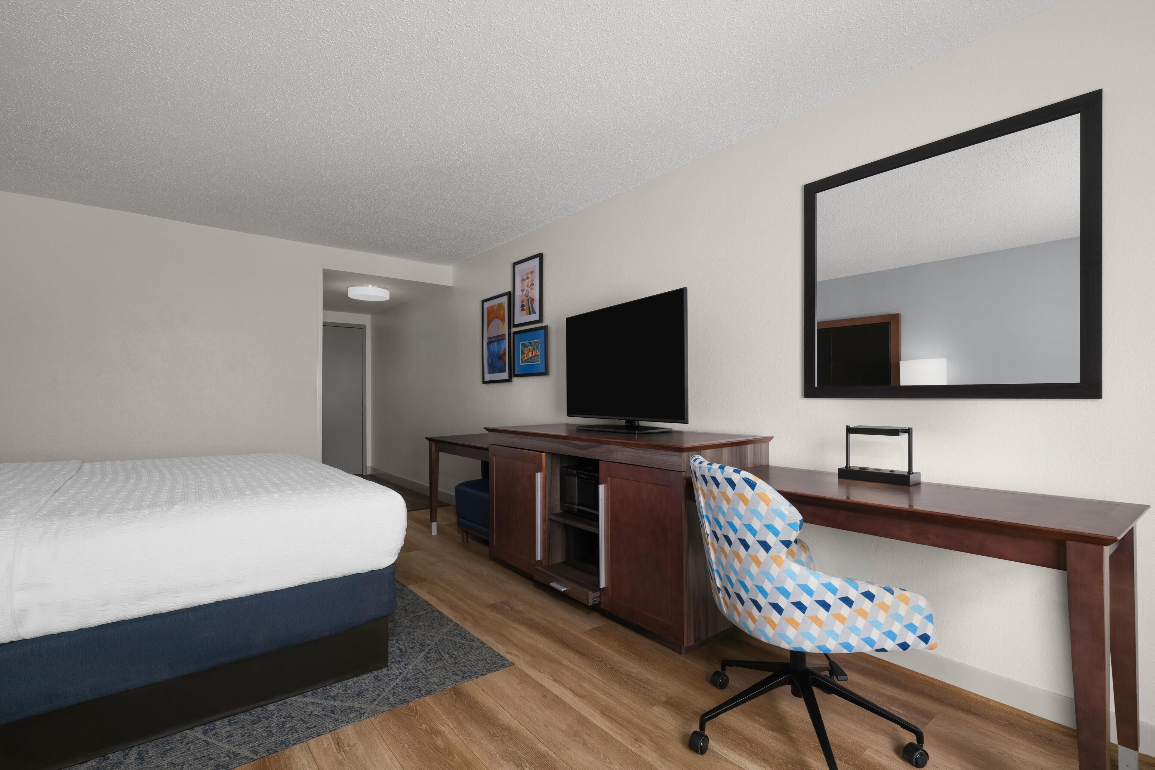 Quarto de hotel com cama, escrivaninha, cadeira, televisão e espelho.