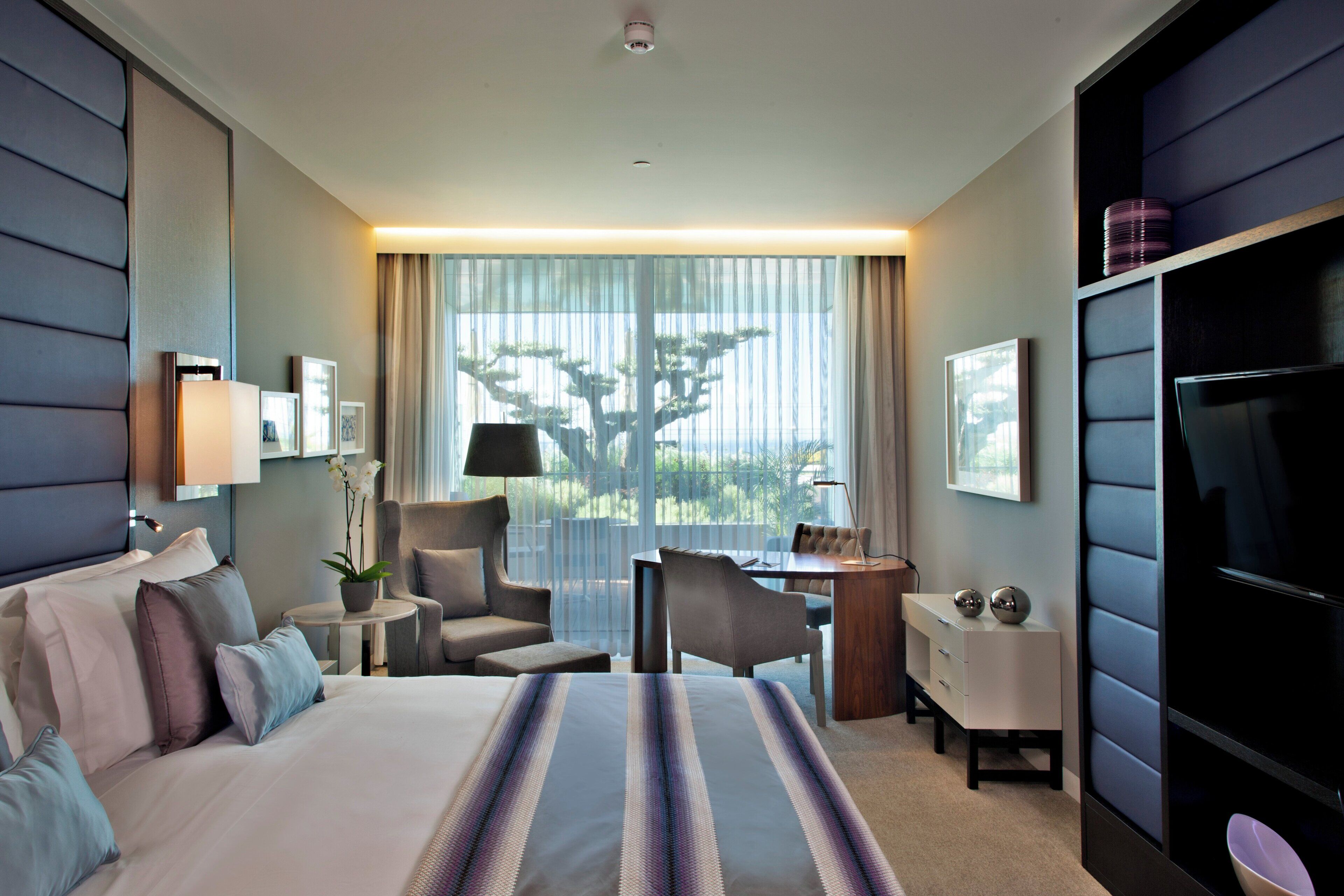 Quarto de hotel com cama, escrivaninha, cadeira, televisão e janela com cortinas.