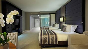 Quarto de hotel com uma cama grande, área de estar, televisão e banheiro visível por uma porta de vidro.