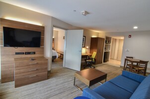 Quarto de hotel moderno com televisão de tela plana fixada em um painel de madeira, cômoda de madeira com gavetas, sofá azul, pequena mesa de madeira e área de cozinha compacta ao fundo.