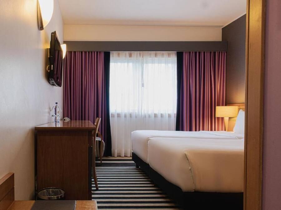 Quarto de hotel com cama, escrivaninha, cadeira, televisão e janela com cortinas.