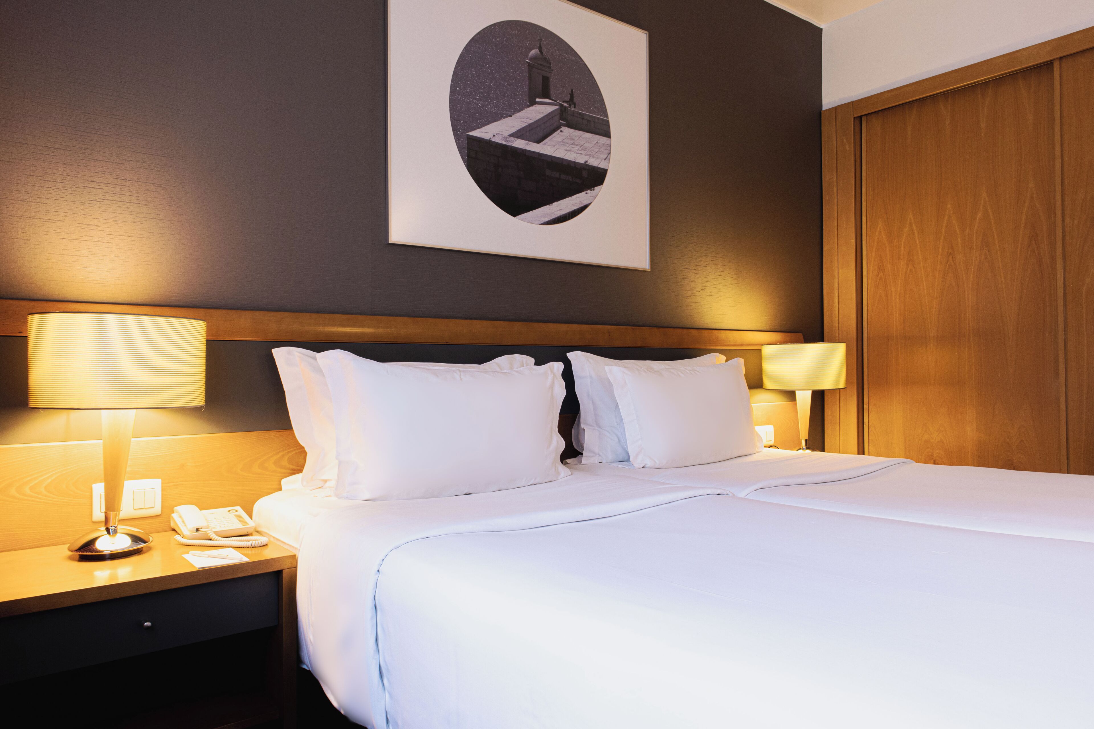 Quarto de hotel com uma cama, duas luminárias de cabeceira, mesa de cabeceira com telefone e um quadro na parede.