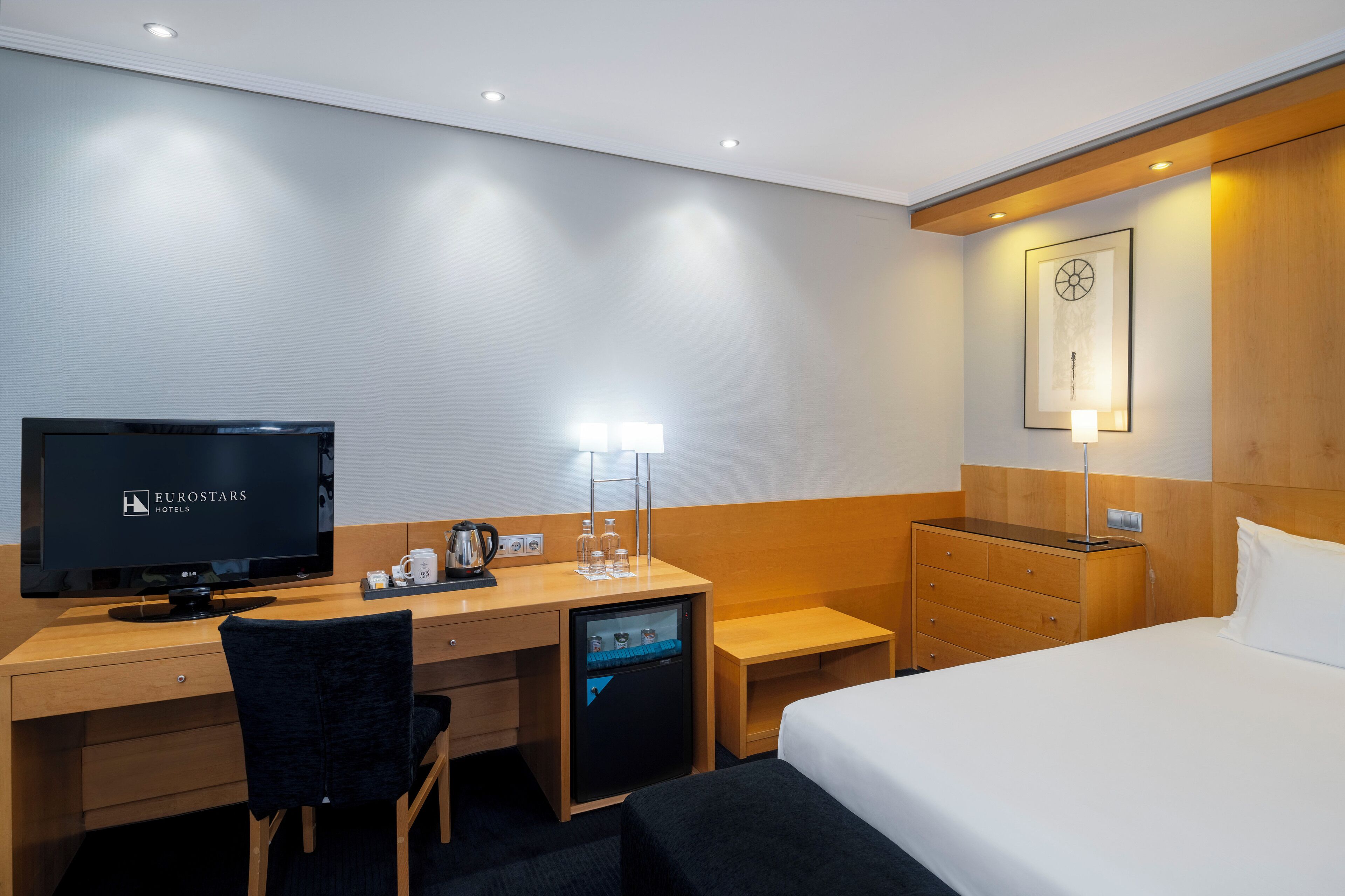 Quarto de hotel com cama, escrivaninha com televisão, cadeira, cómoda e mesa de cabeceira com abajur.