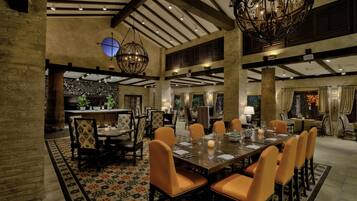 Uma ampla sala de jantar com uma mesa comprida posta para vários convidados, cercada por cadeiras confortáveis. O espaço possui vigas de madeira aparente, um grande candelabro e um tapete estampado.