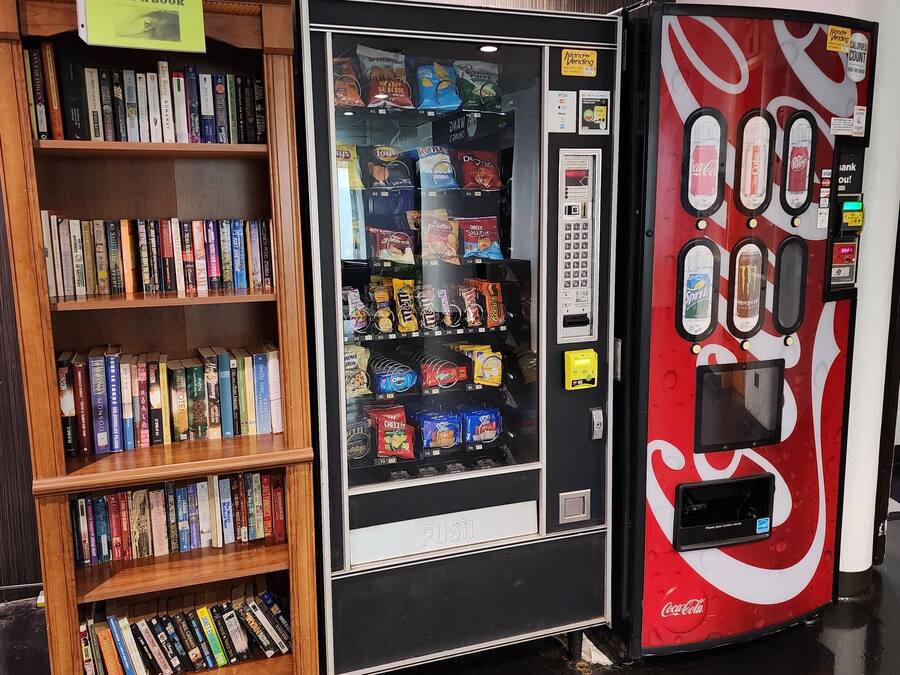 Máquina automática com lanches e distribuidora de Coca-Cola, e uma estante com diversos livros.