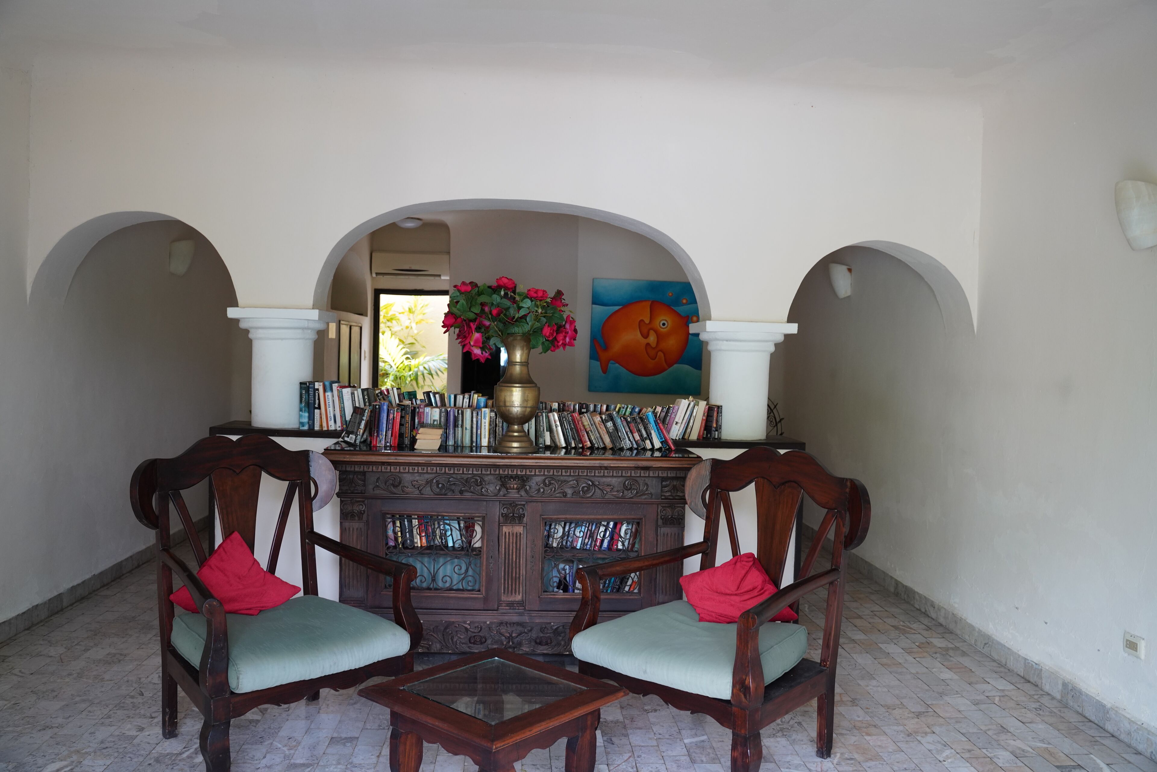 Uma área de leitura com duas cadeiras de madeira, uma mesa de centro de vidro e uma estante repleta de livros e um vaso de flores.
