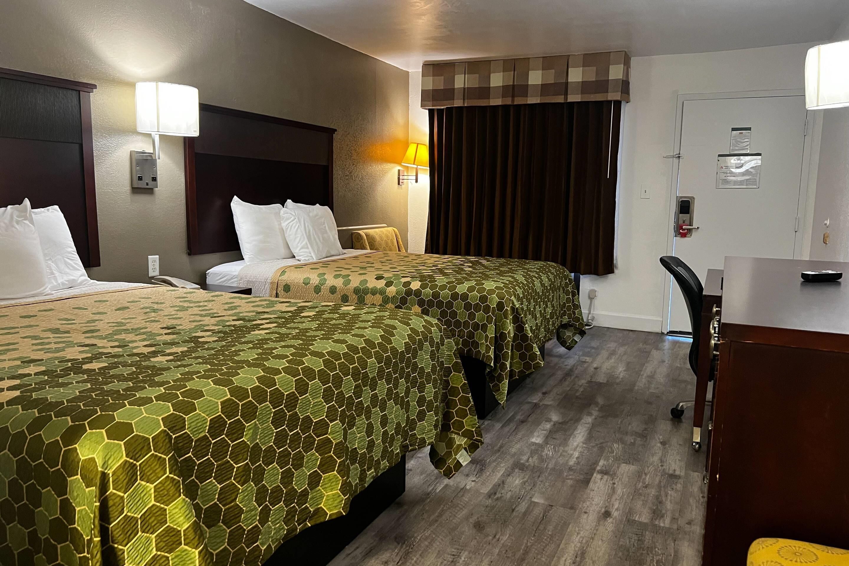 Quarto de hotel com duas camas, uma escrivaninha, uma cadeira e janela com cortinas.