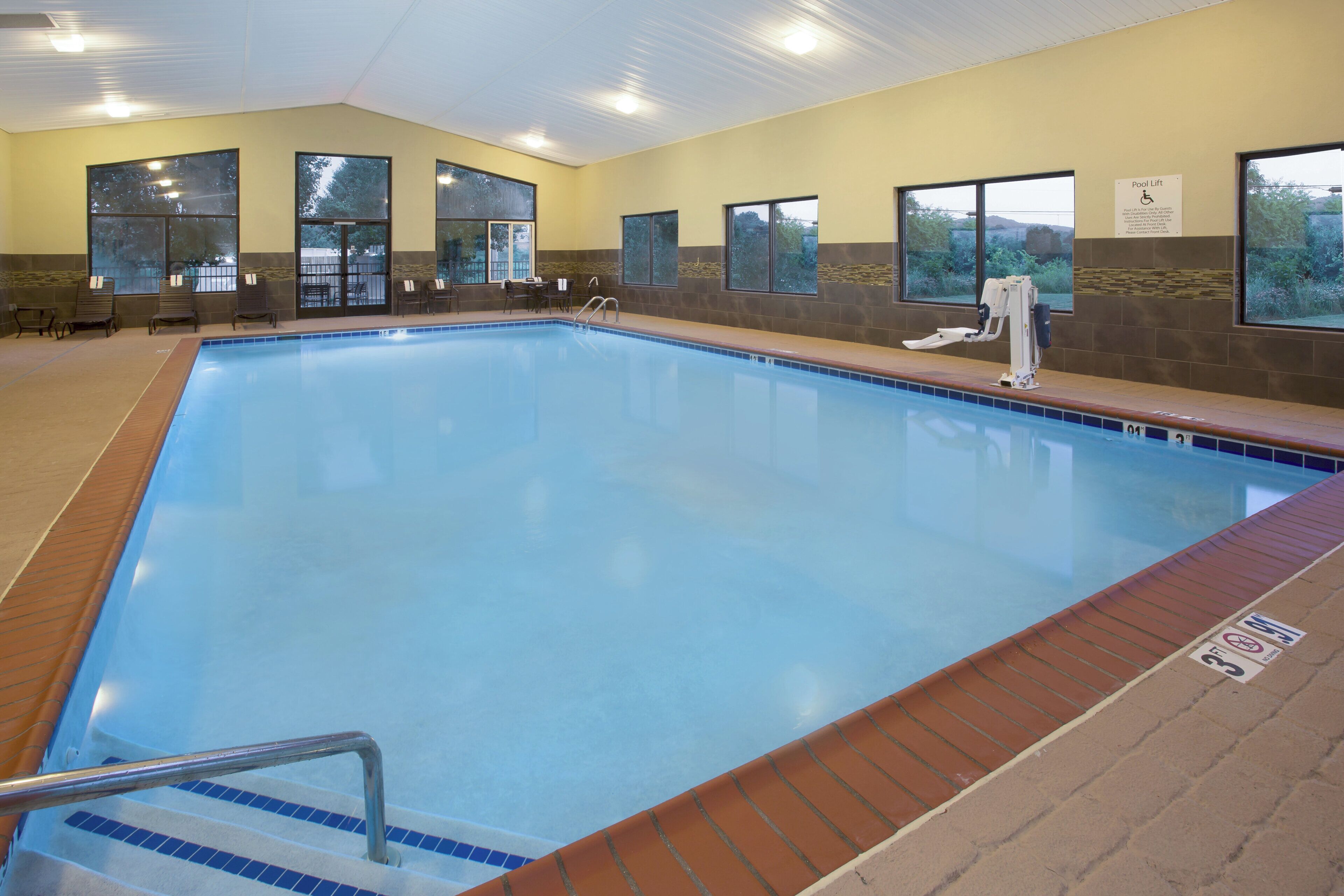 Piscina com elevador de piscina, cadeiras de descanso e vista para o exterior através de grandes janelas.