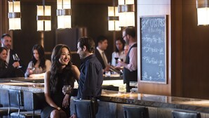 Uma área de bar com cardápio em lousa listando ostras e outros itens, banquetas e luminárias pendentes.