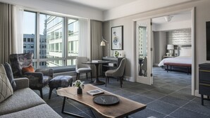 Quarto de hotel moderno com sofá, poltrona, mesa de centro e escrivaninha com luminária. Possui uma cama com cabeceira branca e vista da cidade através de grandes janelas.