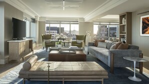Uma sala de estar moderna com sofá, mesa de centro e televisão. Há cadeiras, uma estante e uma grande janela com vista para edifícios.