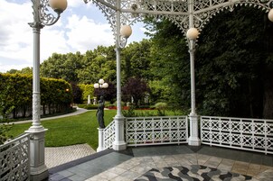Um coreto com grades e colunas brancas ricamente decoradas, rodeado por um jardim bem cuidado com sebes e árvores.