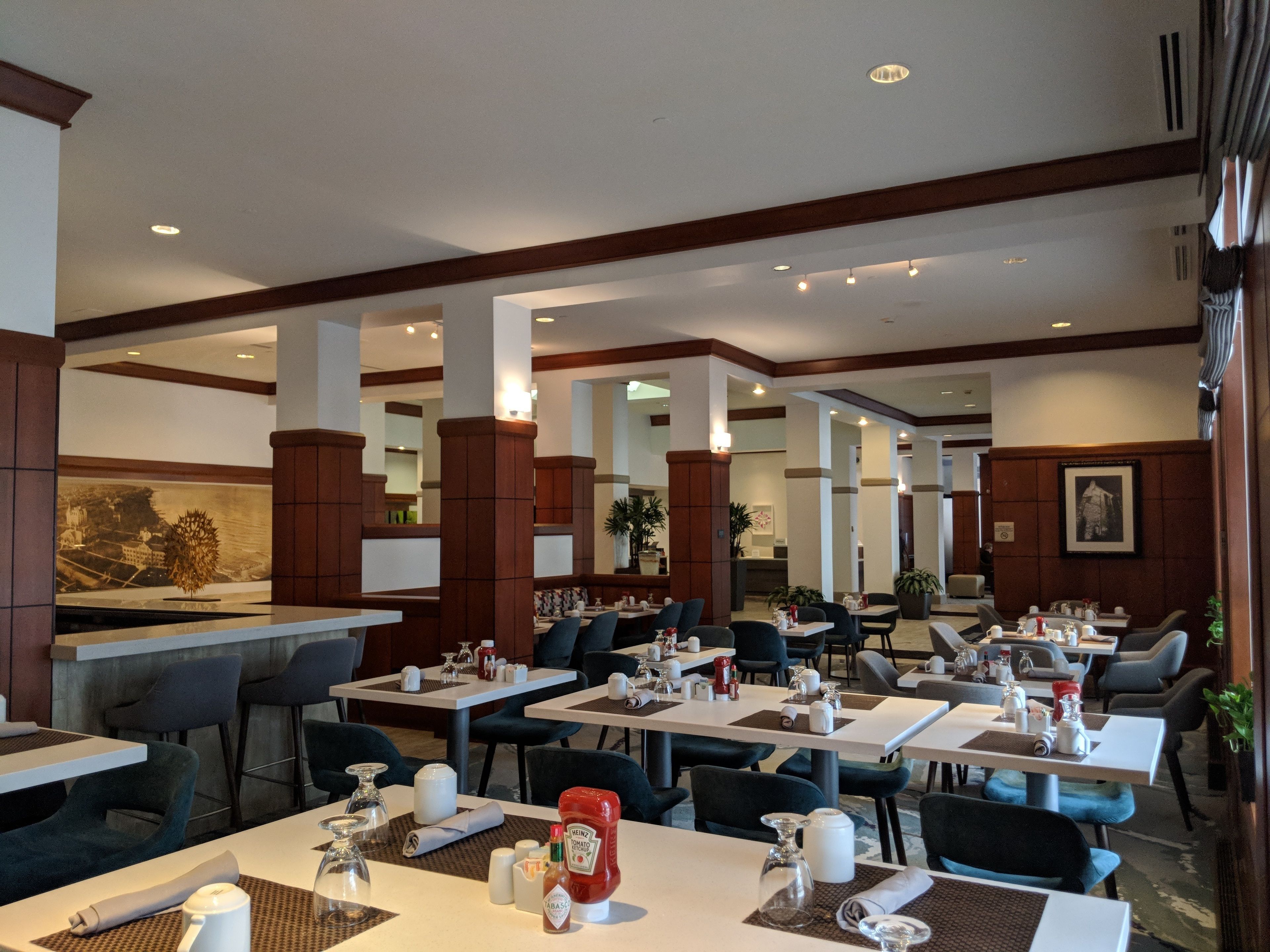 Um restaurante espaçoso com pé-direito alto, diversas áreas de refeições e um bar. As mesas estão dispostas com temperos e copos.