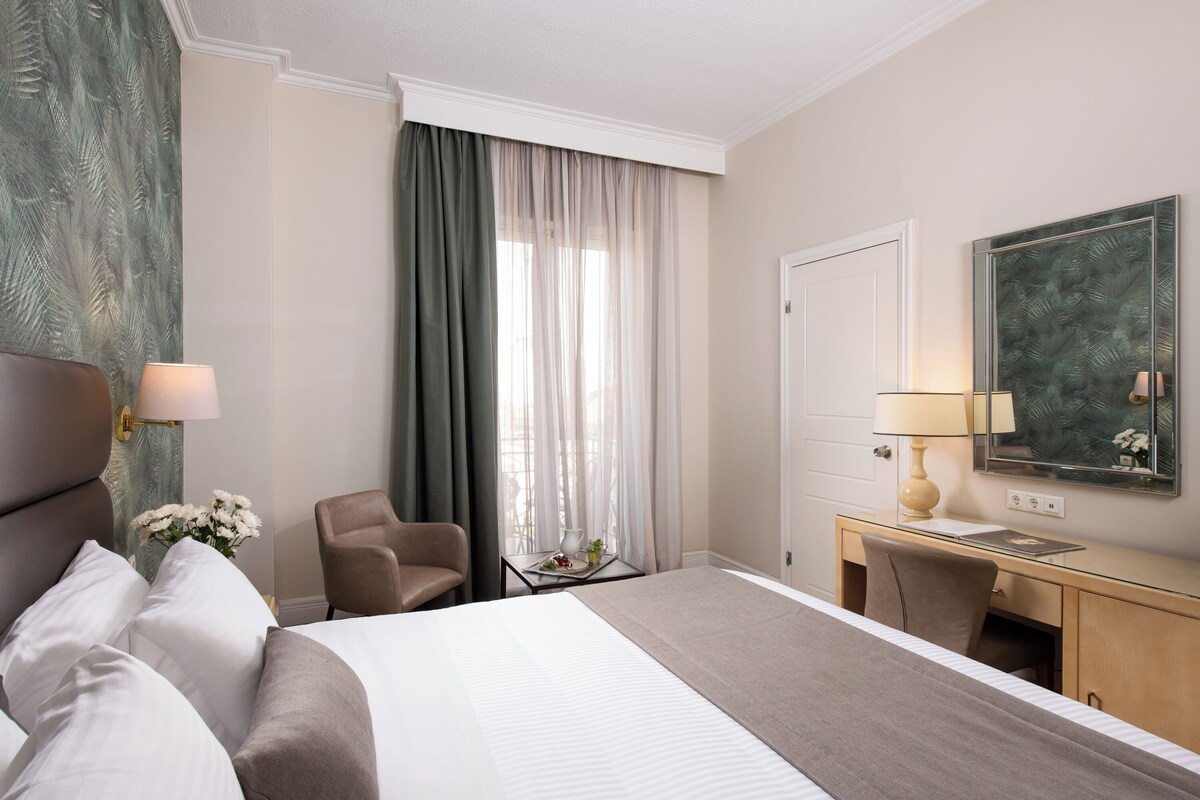 Quarto de hotel com cama, escrivaninha, cadeira, espelho e janela com cortinas.
