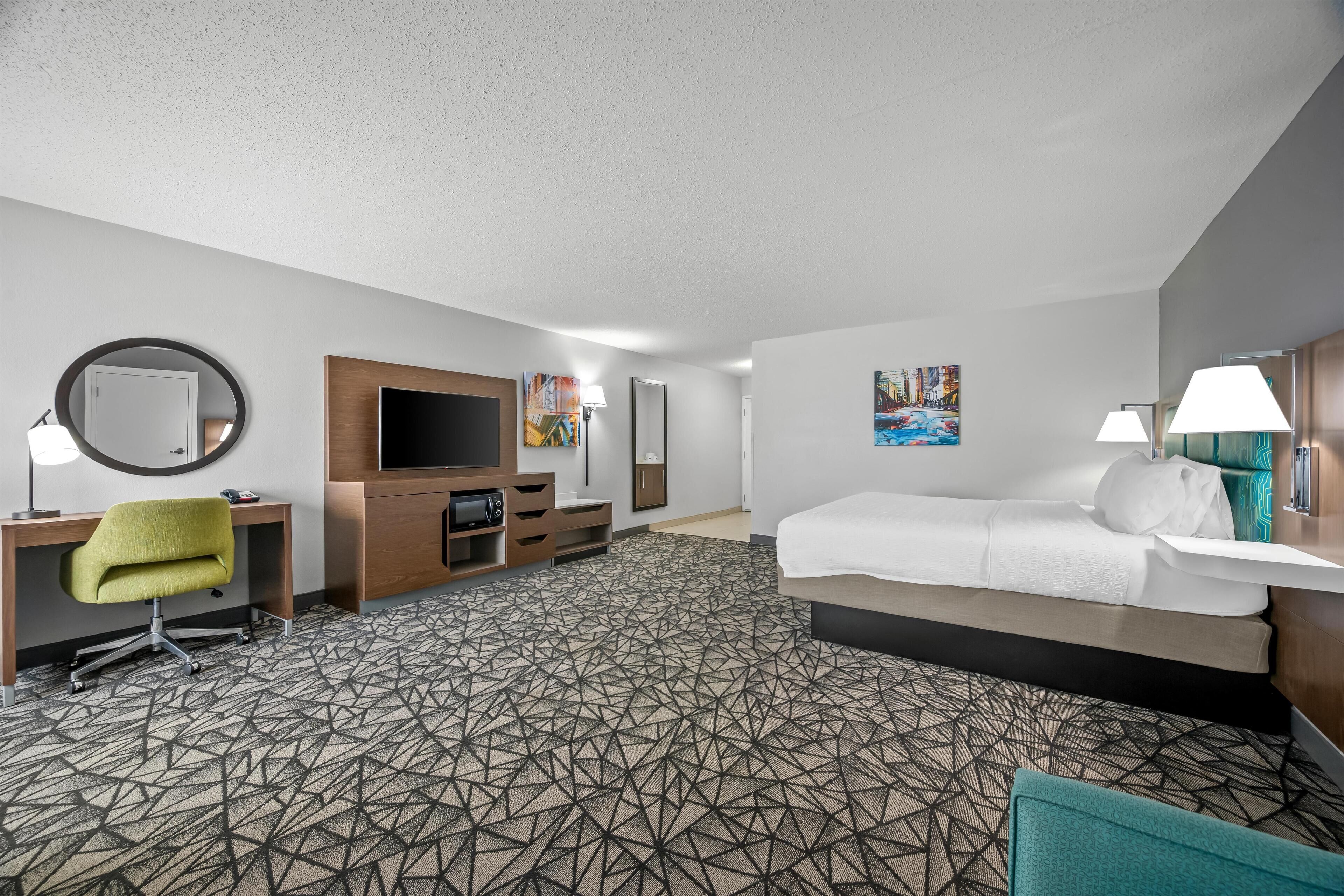 Quarto de hotel com cama, escrivaninha com cadeira, televisão e obras de arte nas paredes.