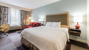 Quarto de hotel com cama grande, escrivaninha, cadeira, sofá vermelho e janela com cortinas.