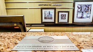 Uma mesa de recepção com uma placa indicando 'welcome to columbus' e quadros nas paredes. Um cartão de fidelidade está sobre o balcão.