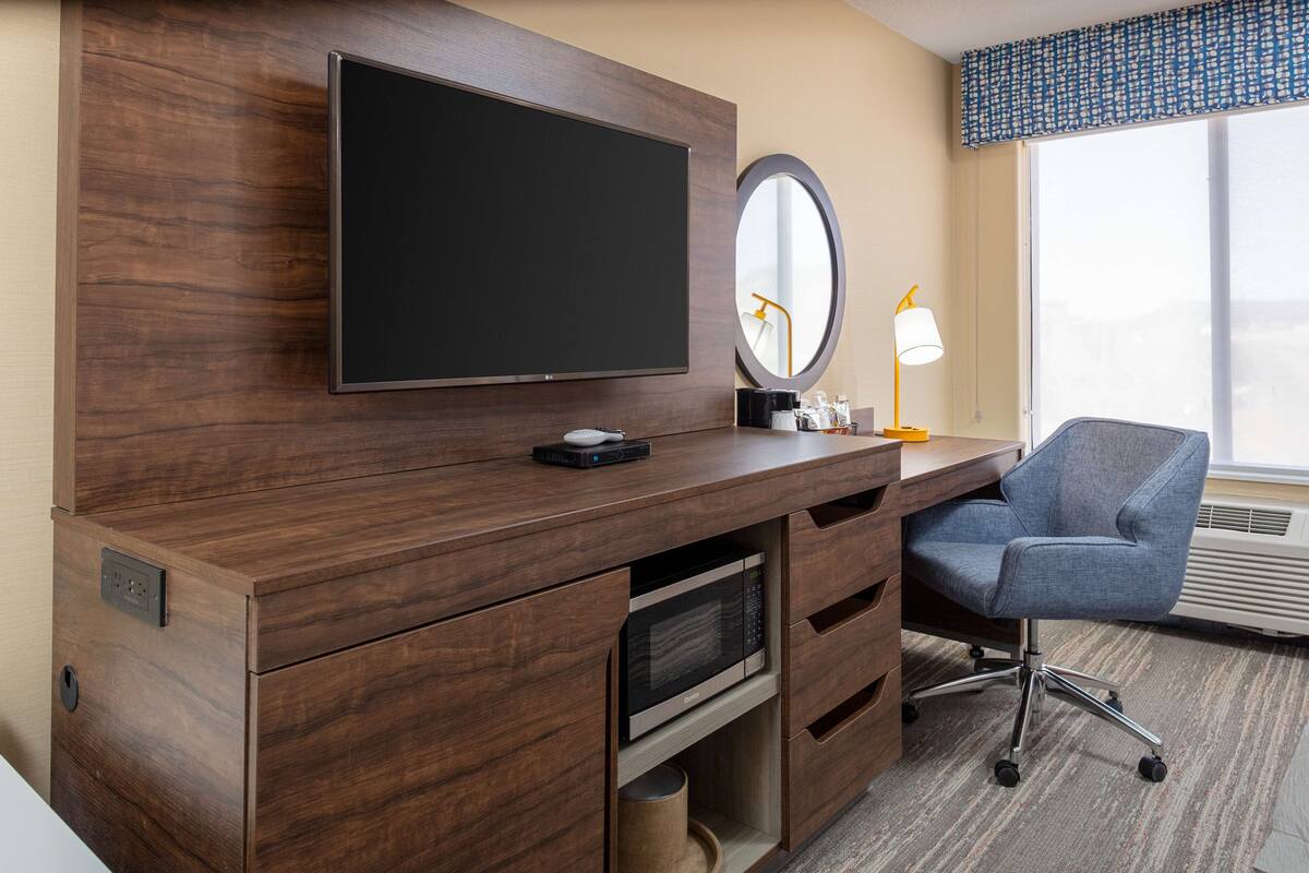 Quarto de hotel com escrivaninha de madeira, televisão de tela plana, micro-ondas, cadeira, espelho e janela com vista.