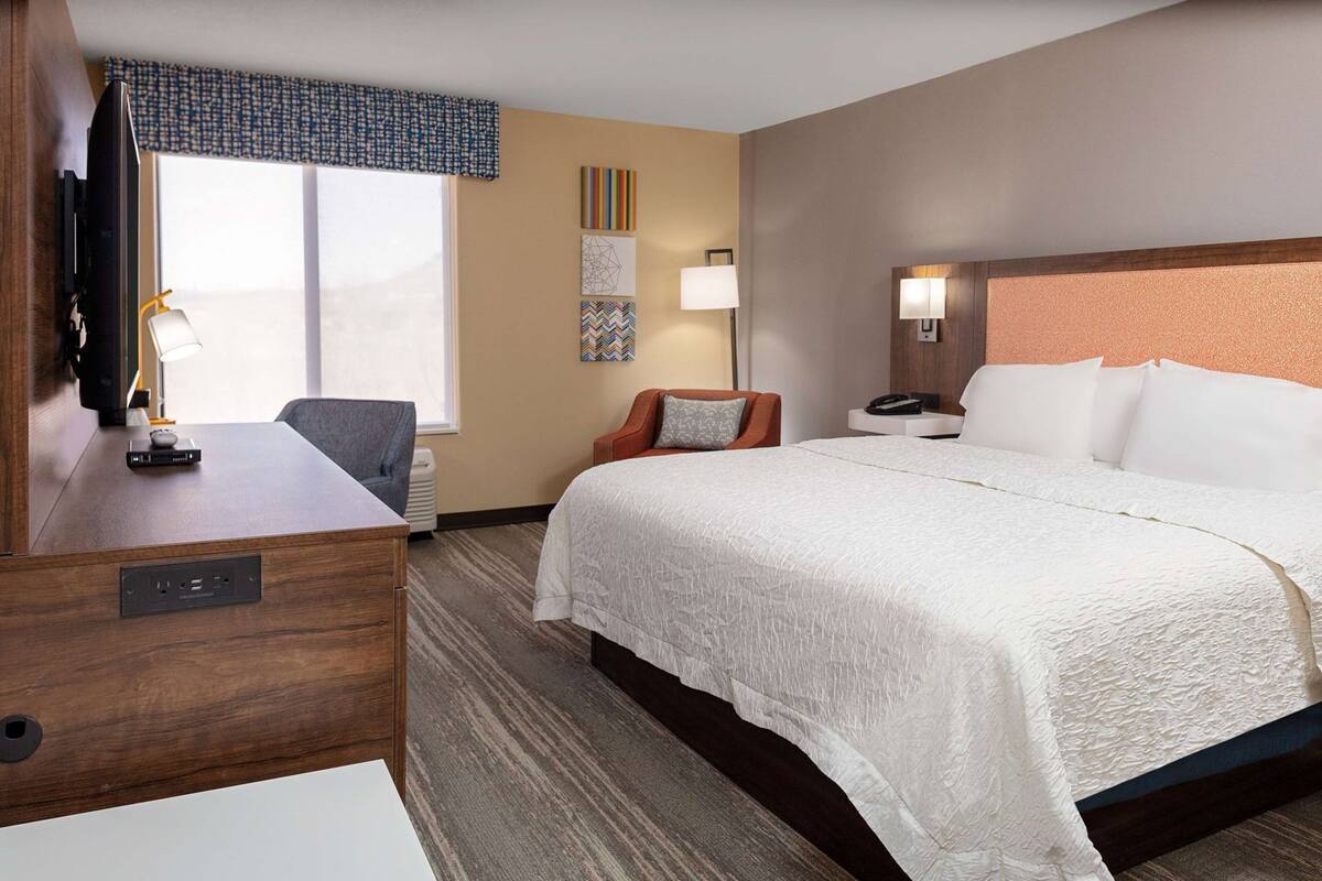 Quarto de hotel com cama, escrivaninha, cadeira e televisão.