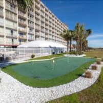 Um hotel com vários andares, área de piscina com gramado verde e palmeiras.