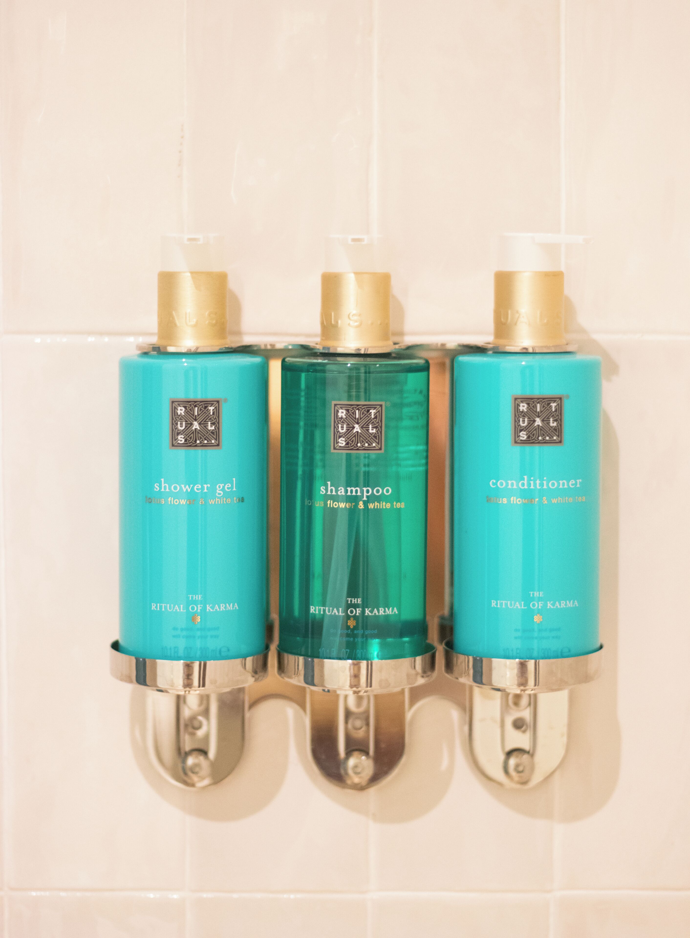 Três produtos Ritual of Karma: gel de banho, champô e condicionador, fixados numa parede.