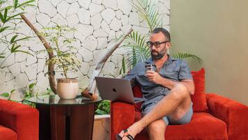 Um homem sentado em um sofá vermelho com um laptop, segurando uma caneca, cercado por plantas e uma parede de pedra.