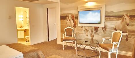 Quarto de hotel com uma televisão grande, duas cadeiras brancas, uma mesa de centro de vidro e chão coberto de carpete.