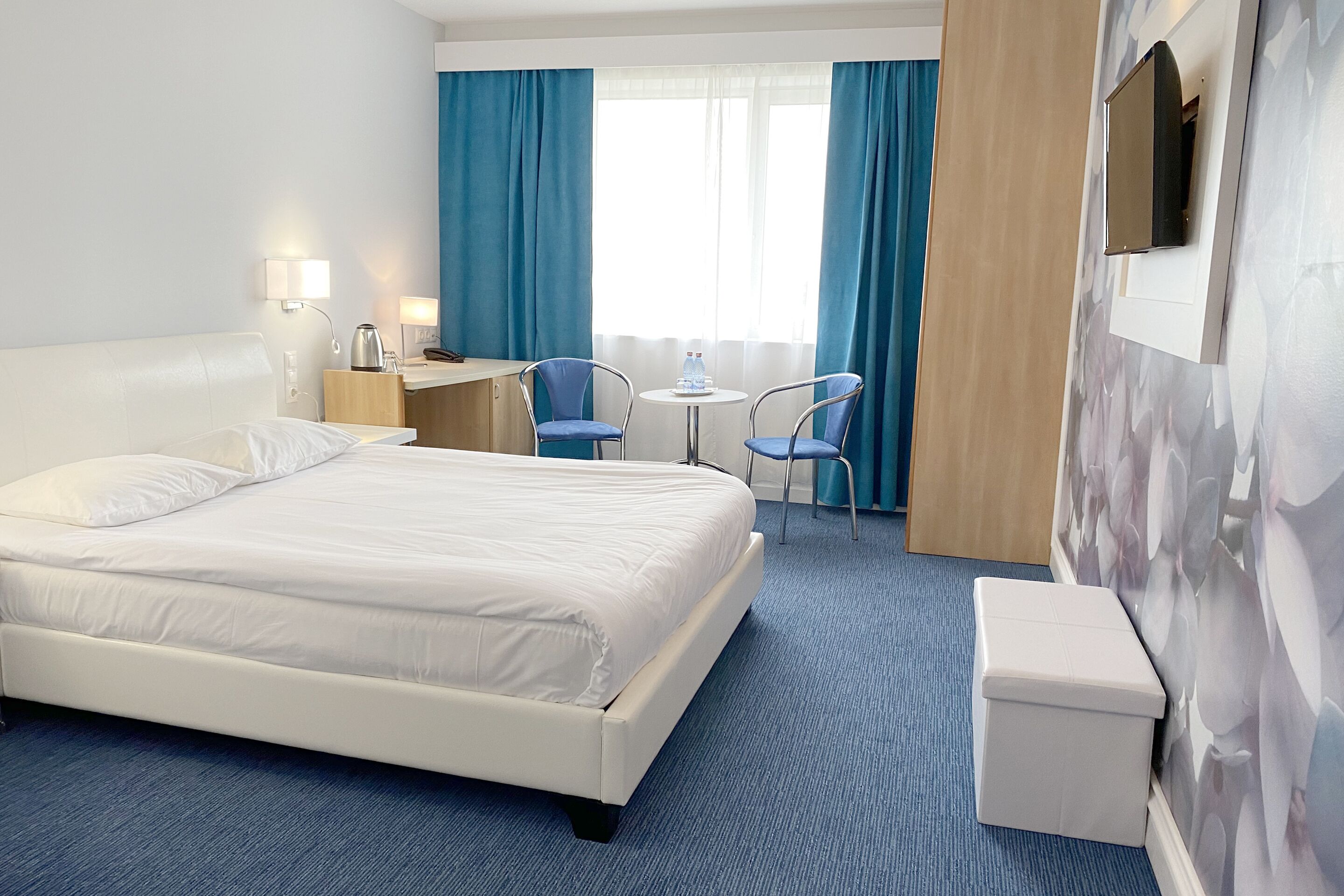 Quarto de hotel com uma cama grande, uma escrivaninha com cadeira, uma mesinha com cadeira e uma televisão fixada na parede.
