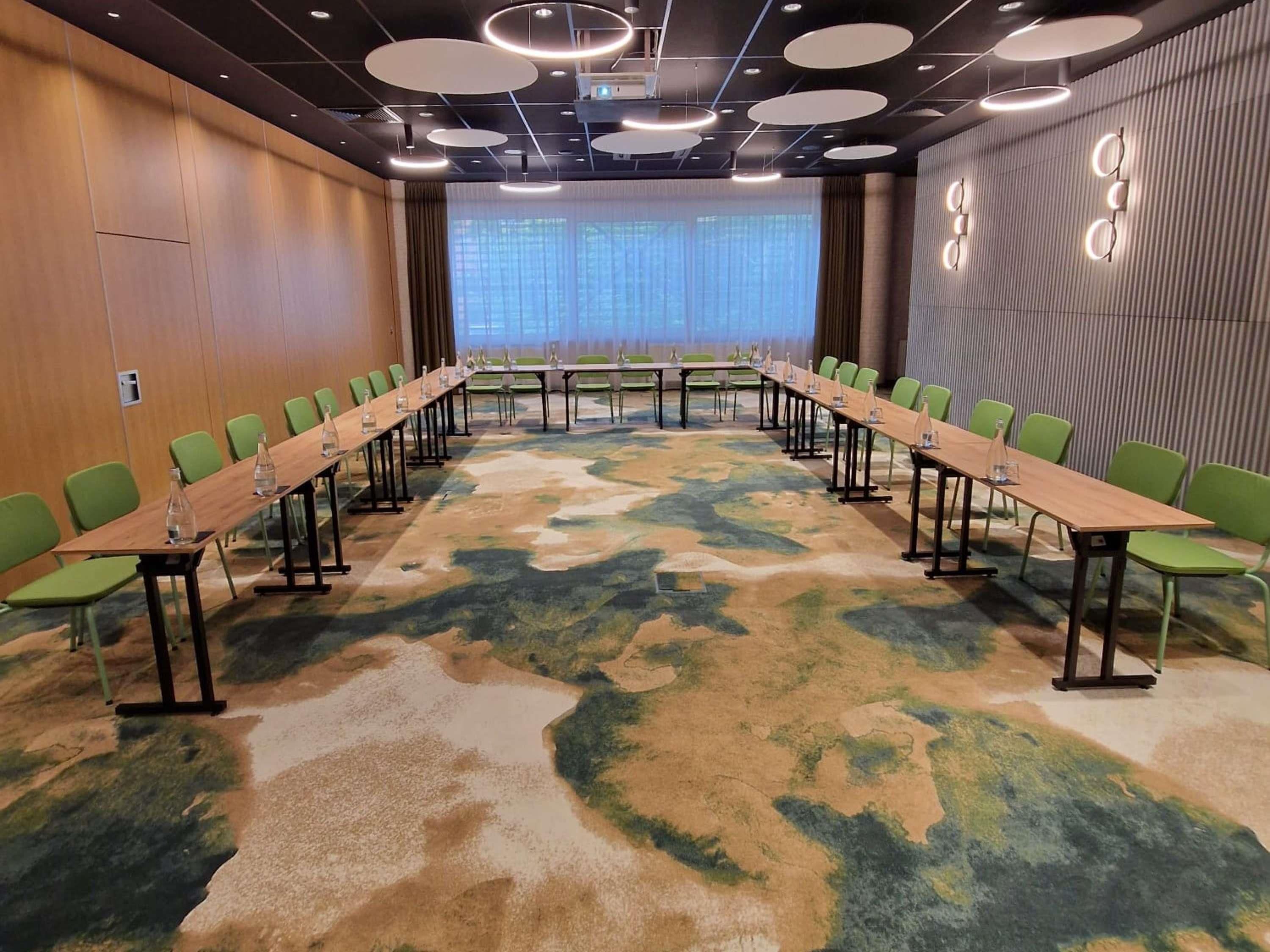 Uma sala de conferências com uma mesa comprida, cadeiras verdes e um tapete estampado.