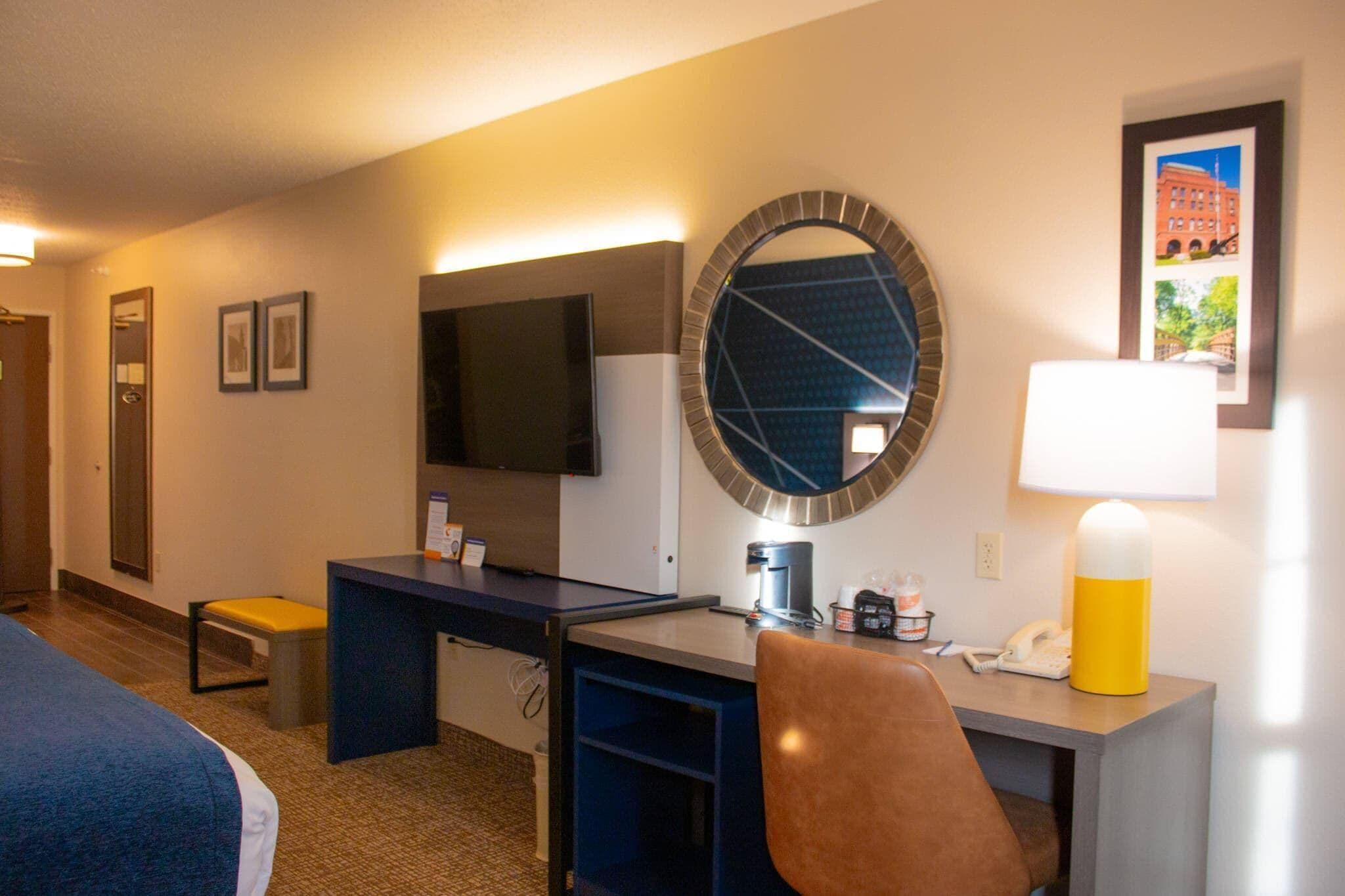Quarto de hotel com cama, escrivaninha e cadeira, televisão, cafeteira, abajur e um quadro na parede.
