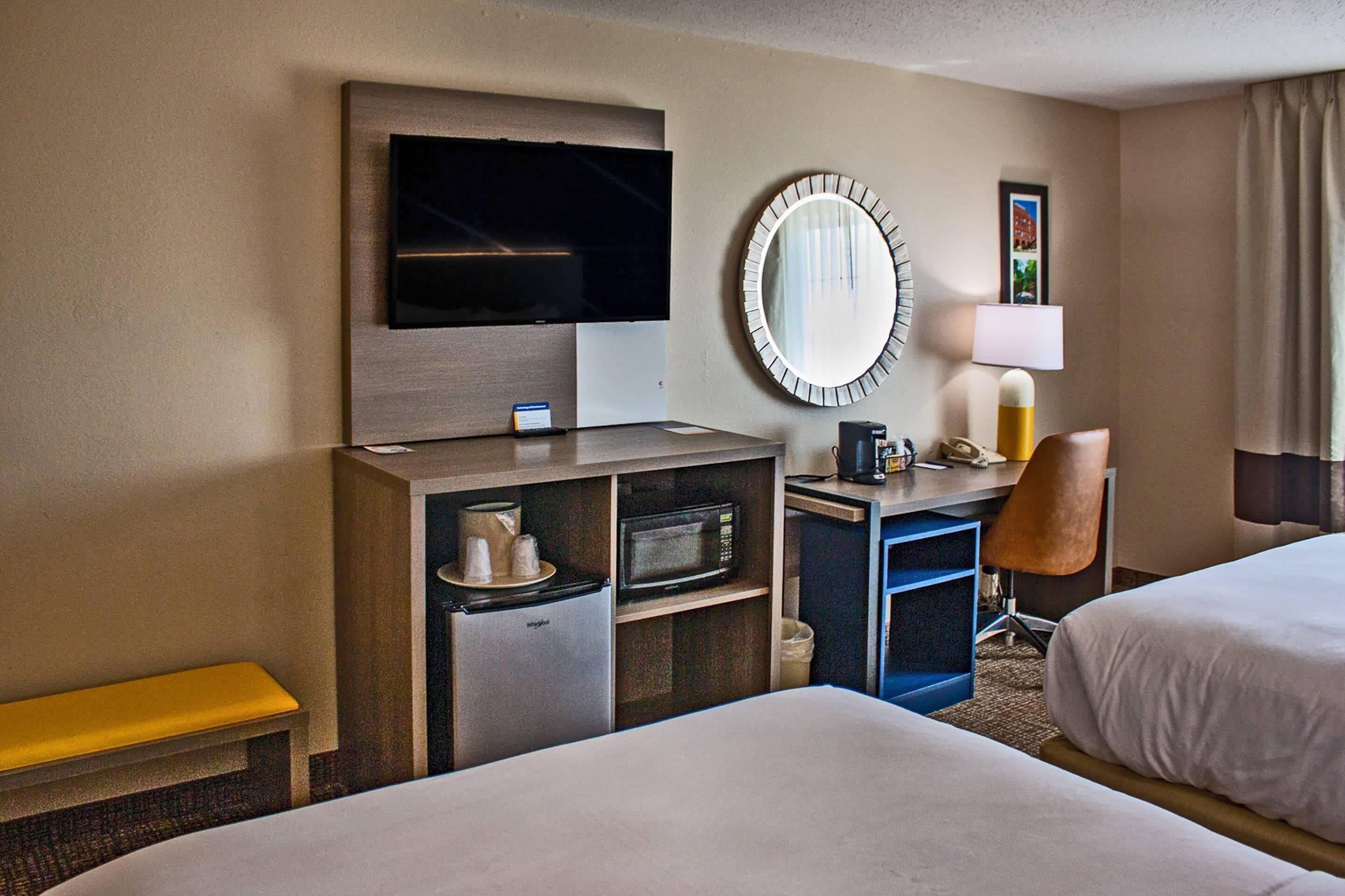 Quarto de hotel com duas camas, TV de tela plana, micro-ondas, cafeteira, espelho e escrivaninha com cadeira.