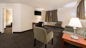 Quarto de hotel com área de estar, televisão fixada na parede, escrivaninha com luminária e janela com cortinas.