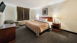 Quarto de hotel com cama, escrivaninha, cadeira, televisão, janela com cortinas e abajur.