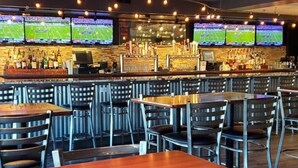 Uma área de bar com várias telas exibindo jogos esportivos, um balcão repleto de garrafas e algumas mesas e cadeiras.